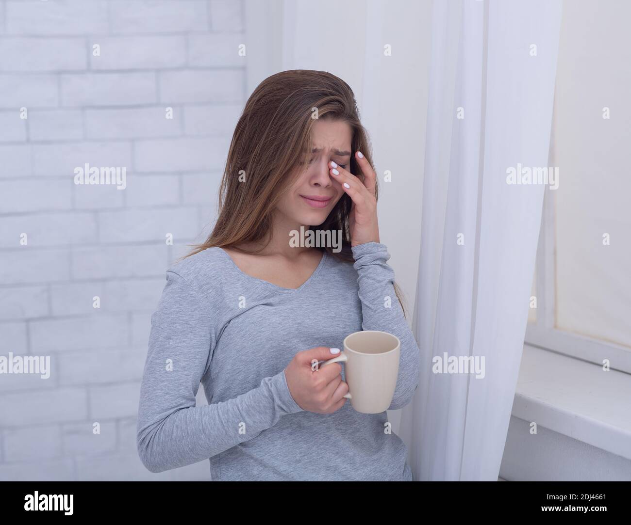 Sad young woman with cup of coffee crying near window, wiping her tears ...