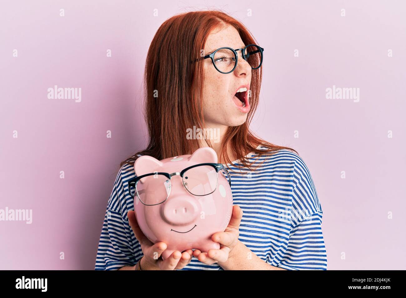Young red head girl holding piggy bank with glasses angry and mad ...