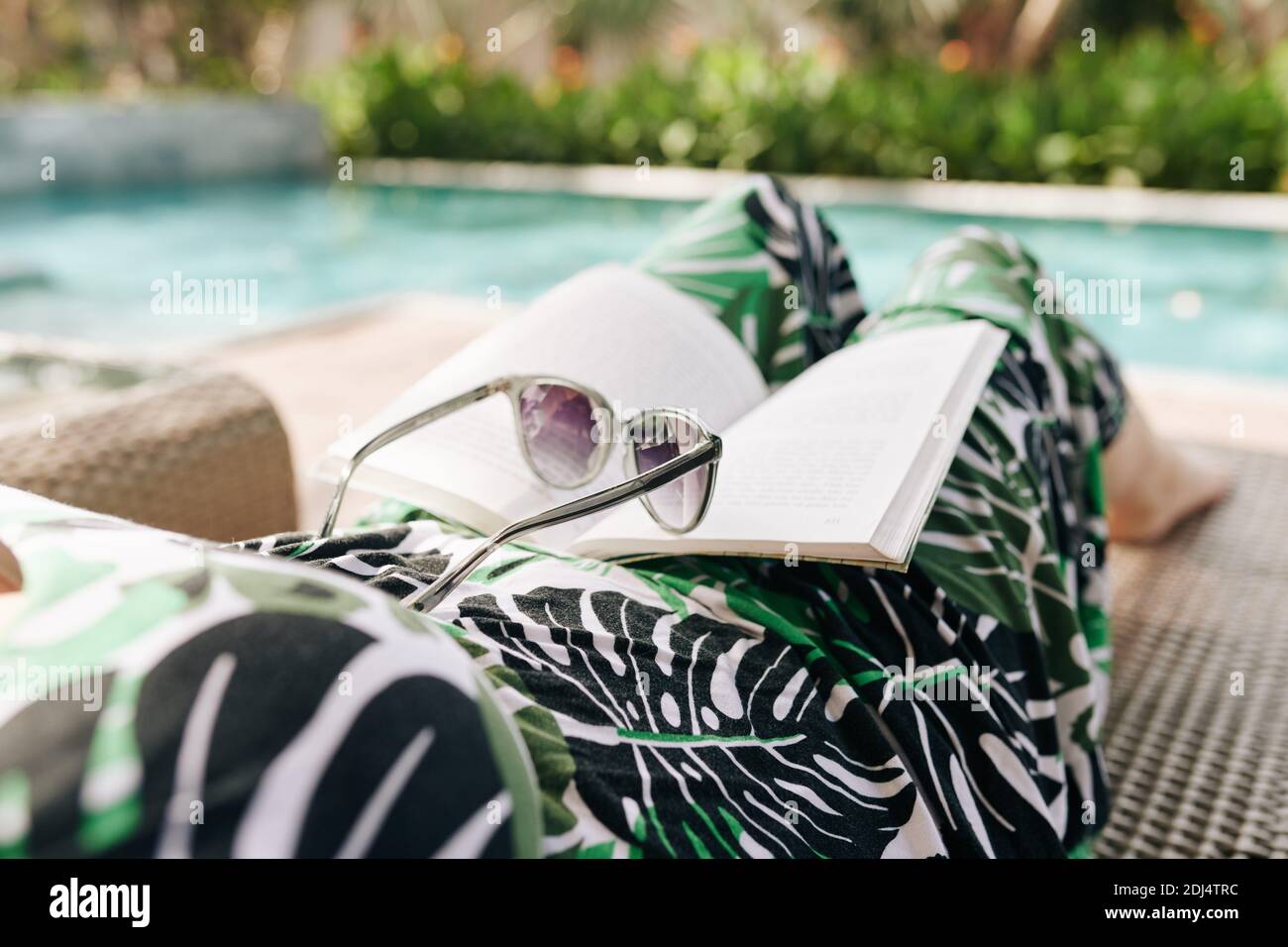 Woman resting by swimming pool with opened book and sunglasses and ...