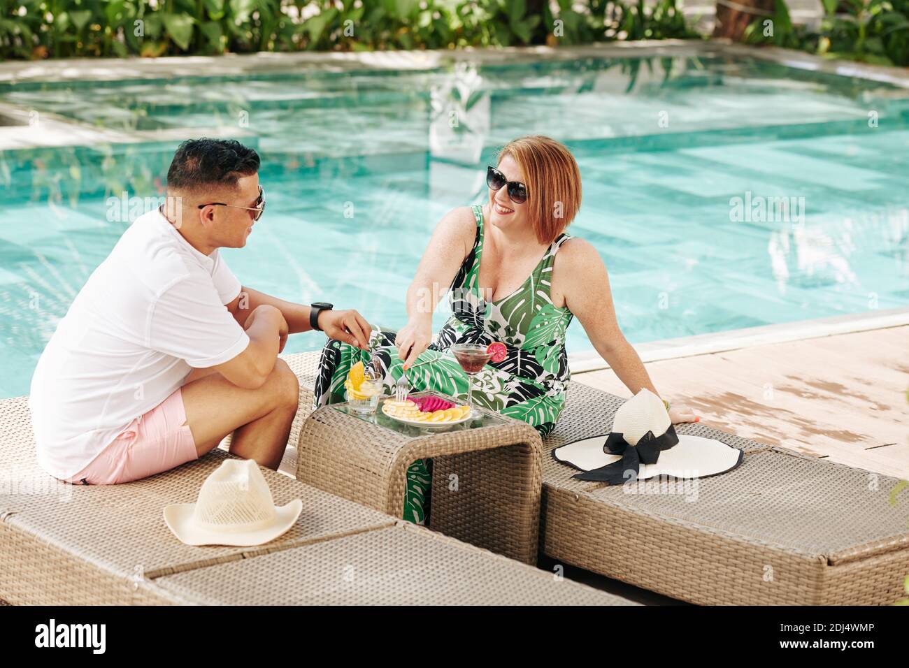 Cheerful couple in love resting by swimming pool, eating cut fruits and ...