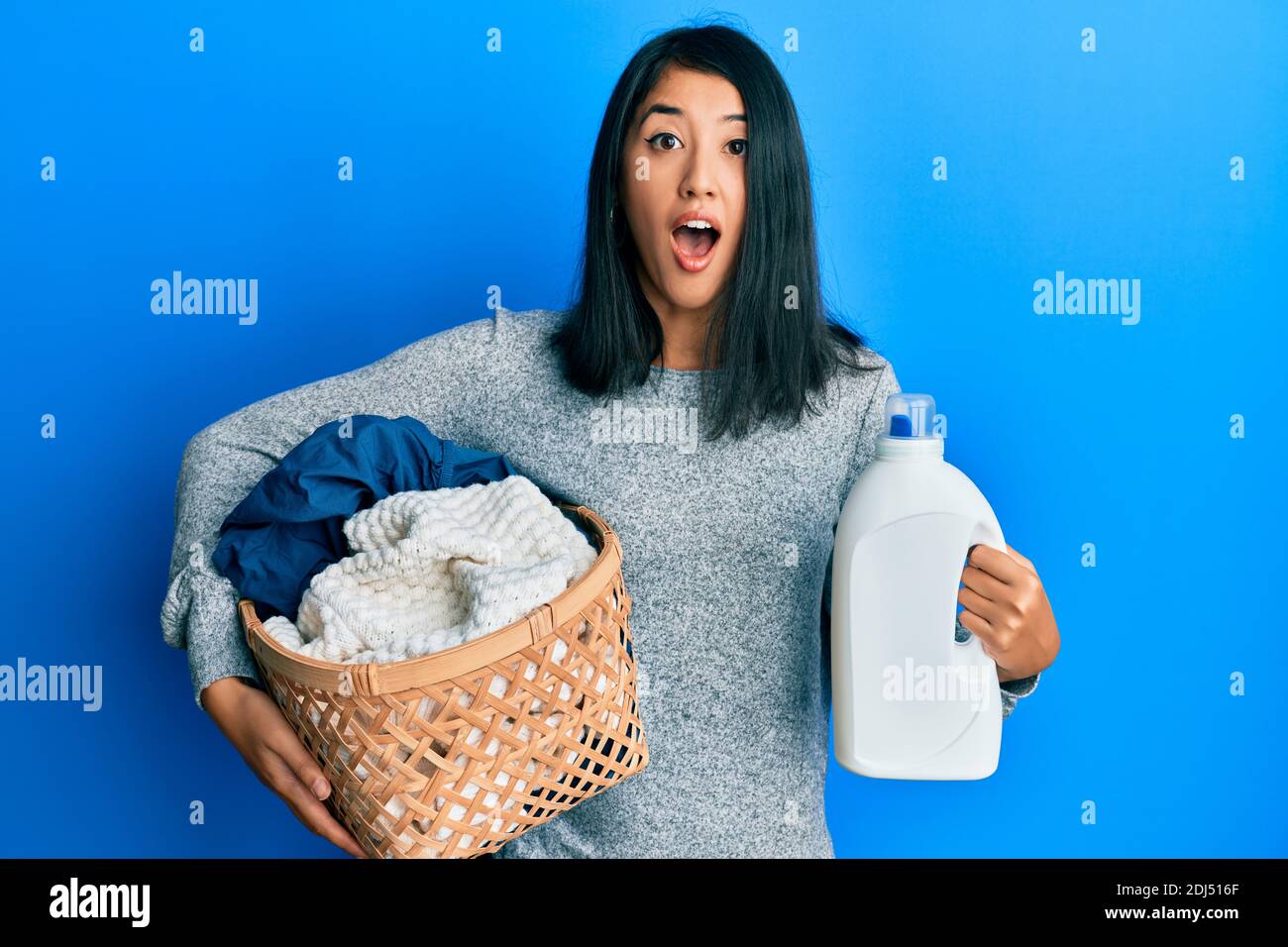 Beautiful asian young woman holding laundry basket and detergent bottle afraid and shocked with ...