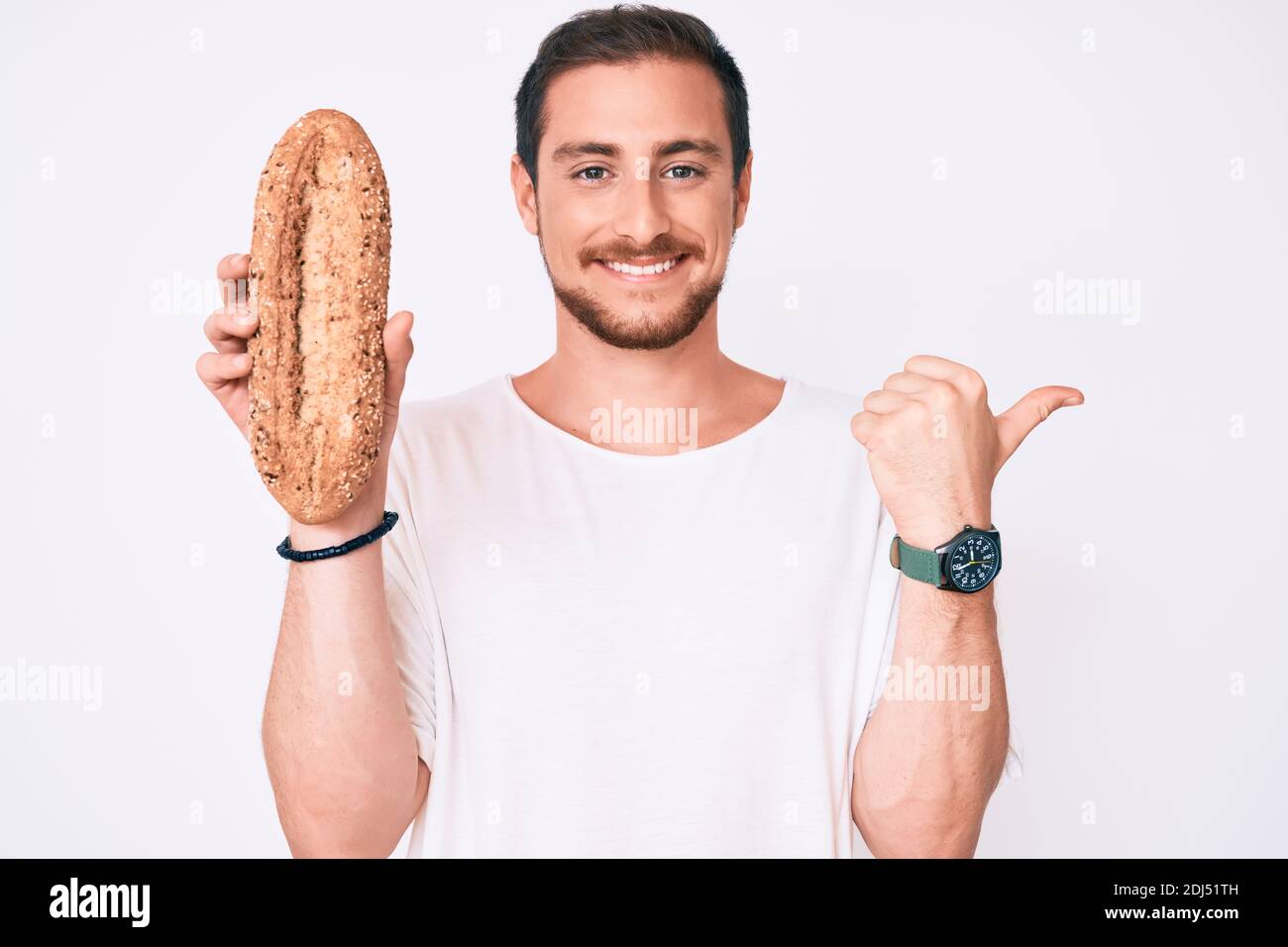Young handsome man holding wholemeal bread pointing thumb up to the ...