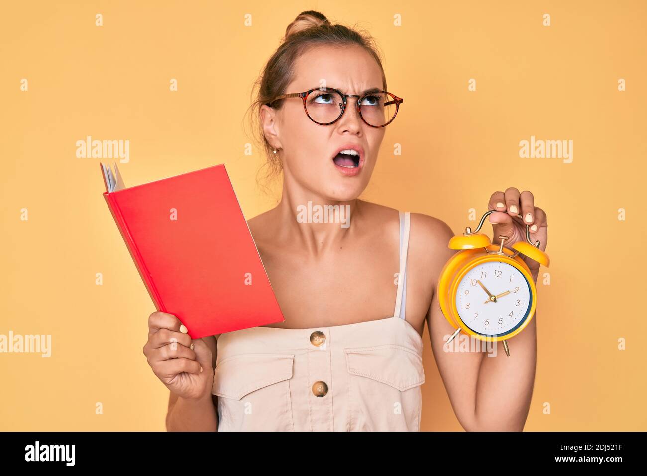 Beautiful caucasian woman holding book and alarm clock angry and mad ...