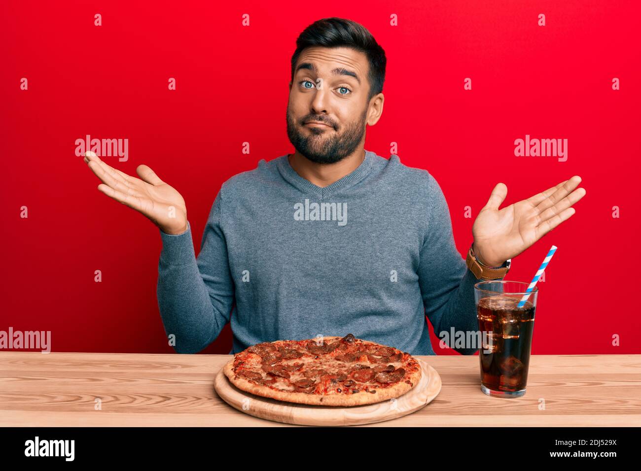 Handsome hispanic man eating tasty pepperoni pizza clueless and ...