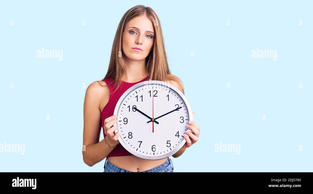 Young beautiful blonde woman holding big clock thinking attitude and ...