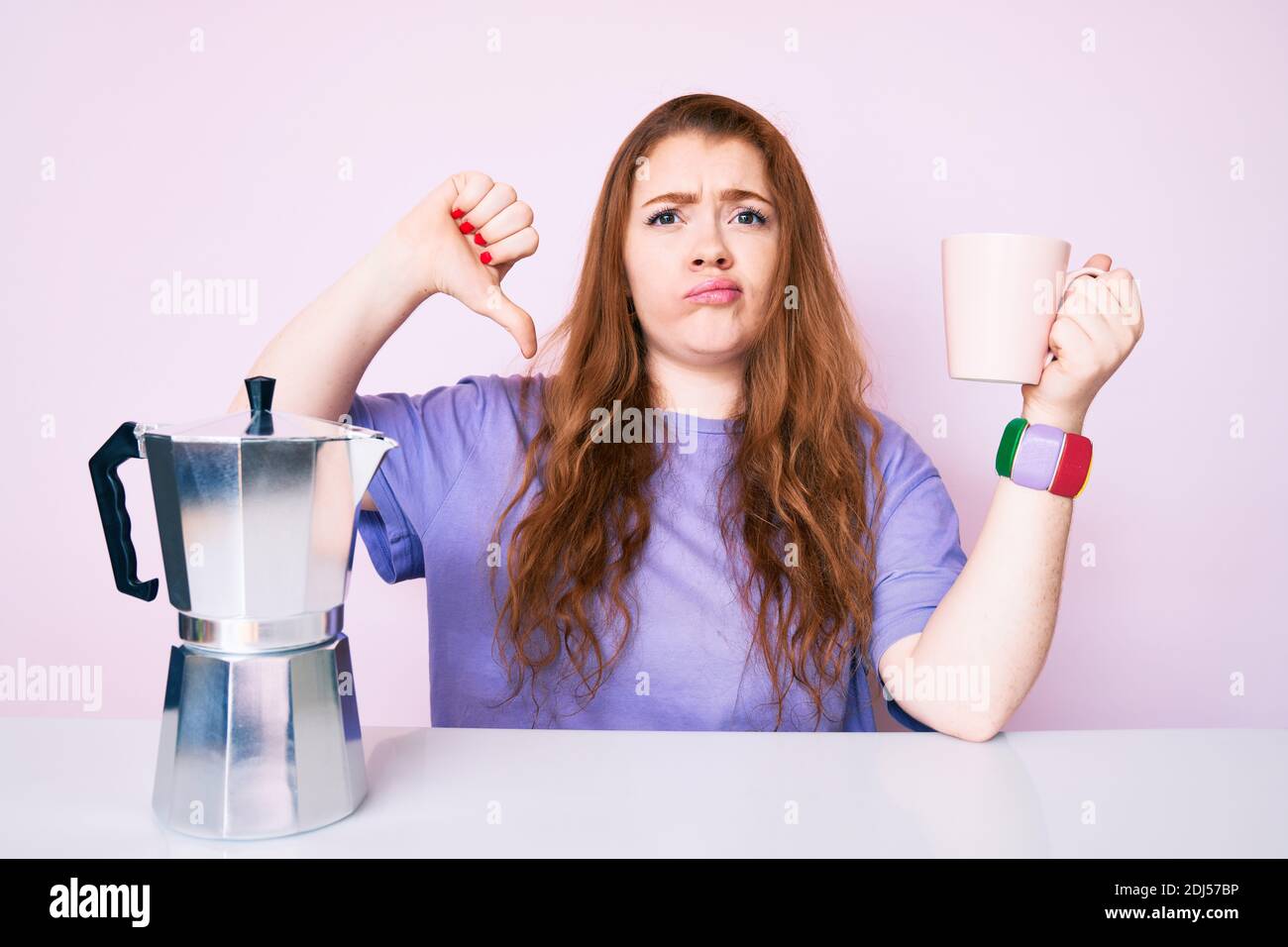 Young redhead woman sitting on the table drinking coffee with angry ...