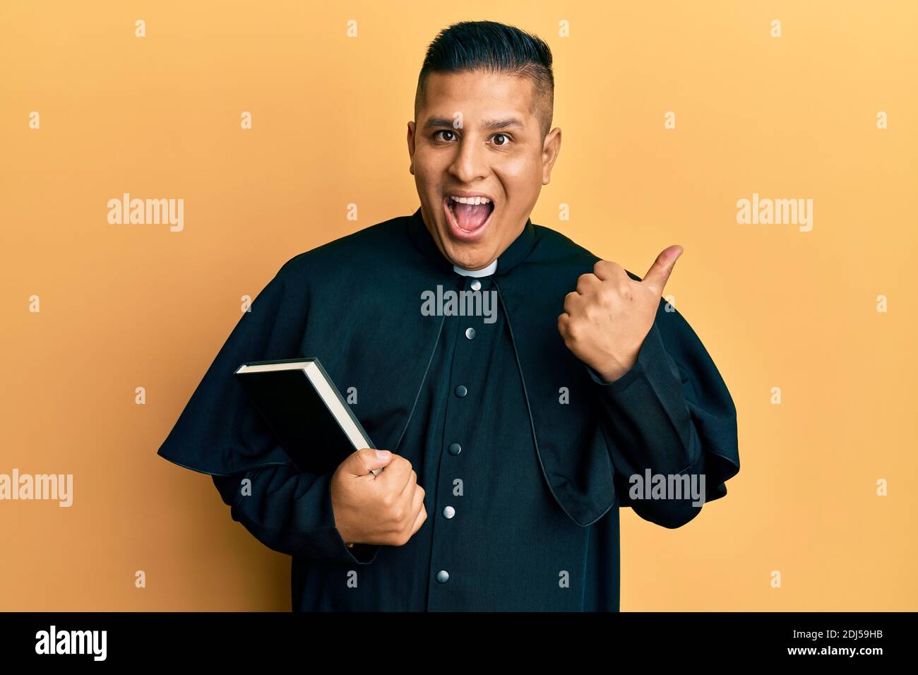 Young latin priest man holding bible pointing thumb up to the side ...
