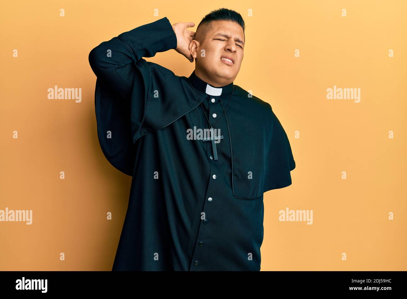 Young latin priest man standing over yellow background suffering of ...