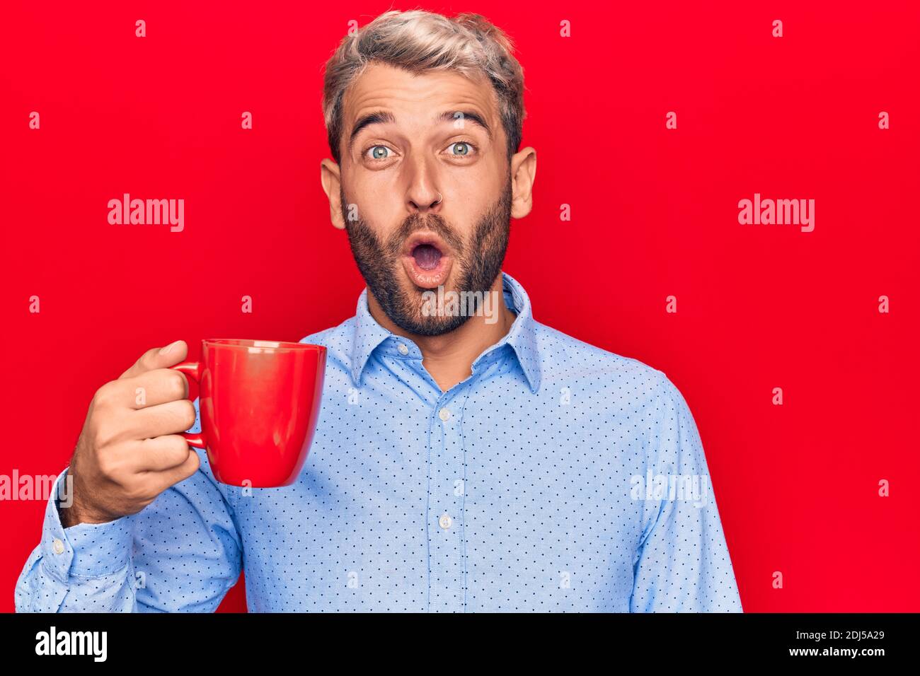Young handsome blond man with beard drinking red mug of coffee over ...