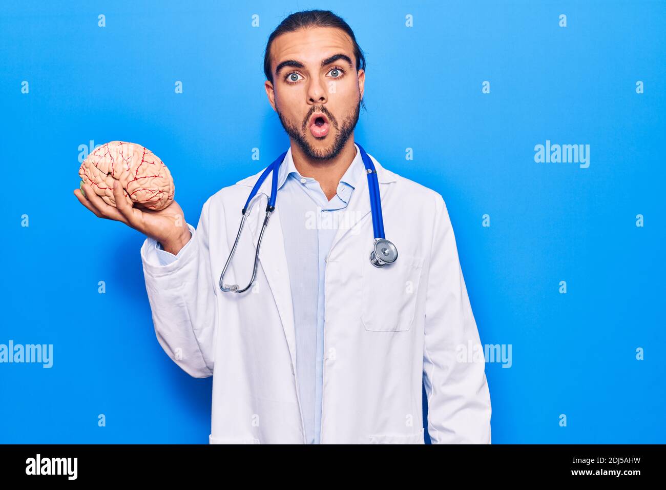 Young handsome man wearing doctor coat holding brain scared and amazed ...