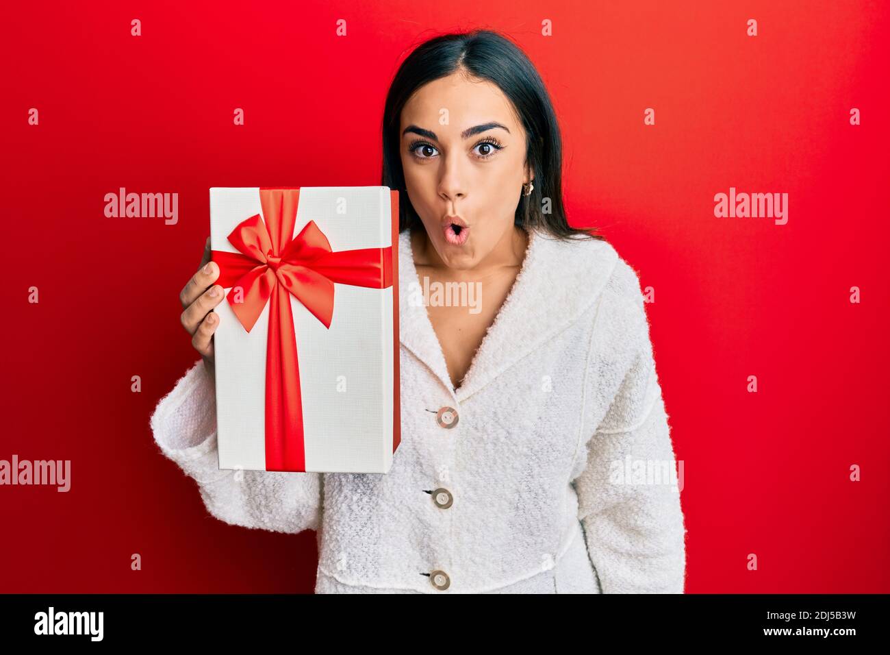 Young brunette woman holding gift scared and amazed with open mouth for ...