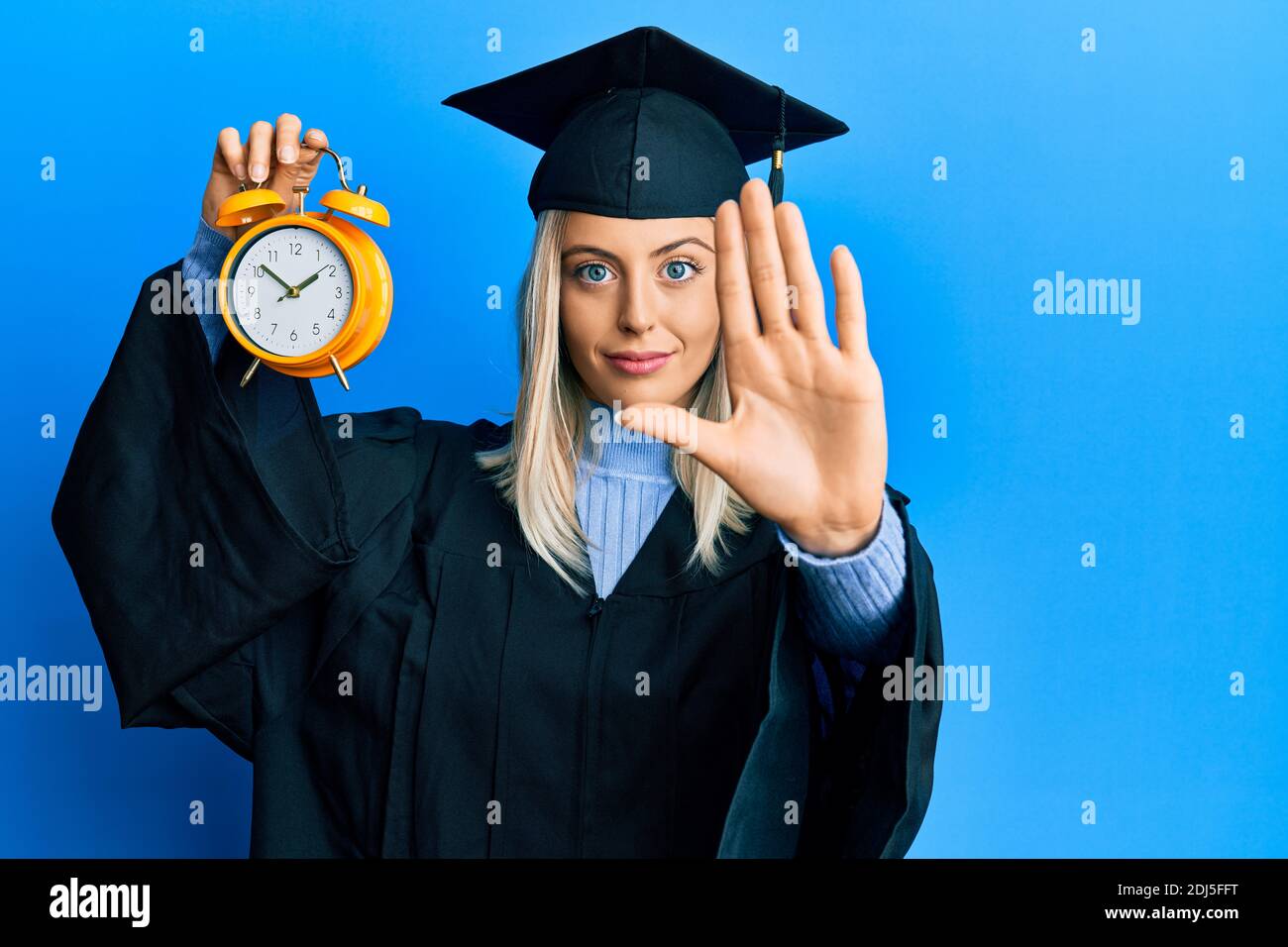 Beautiful blonde woman wearing graduation cap and ceremony robe holding ...