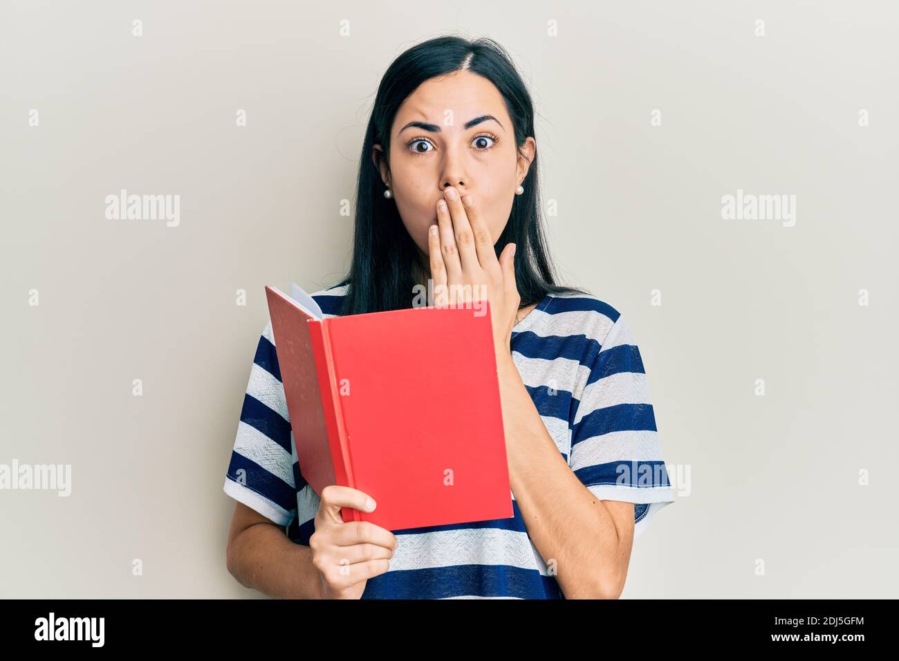 Beautiful young woman reading book covering mouth with hand, shocked ...