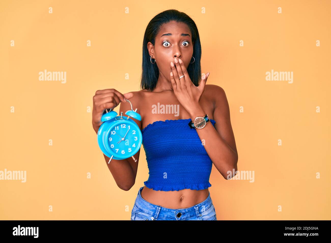 Young african american girl holding alarm clock covering mouth with ...