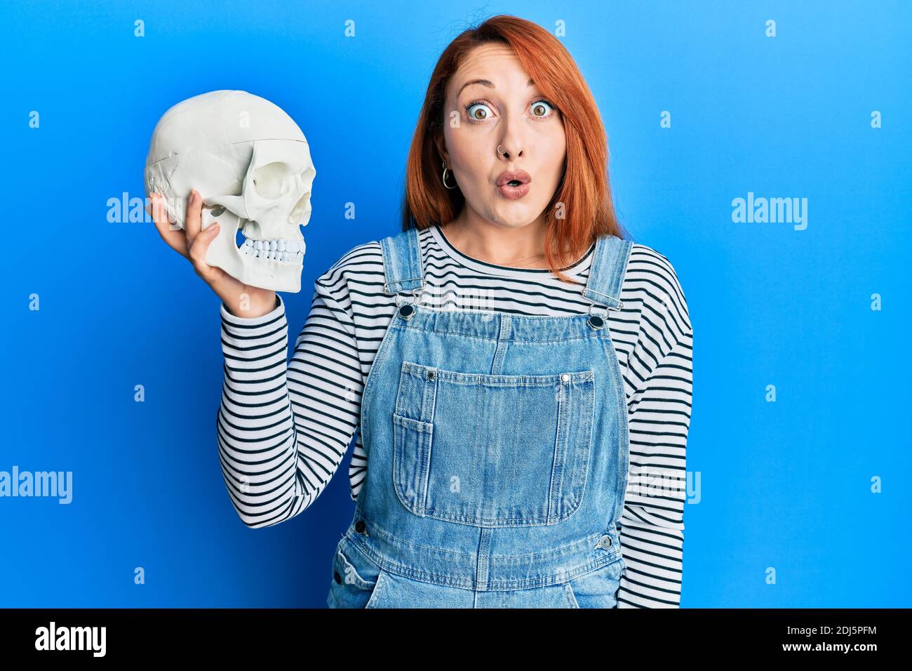 Beautiful redhead woman holding human skull scared and amazed with open ...