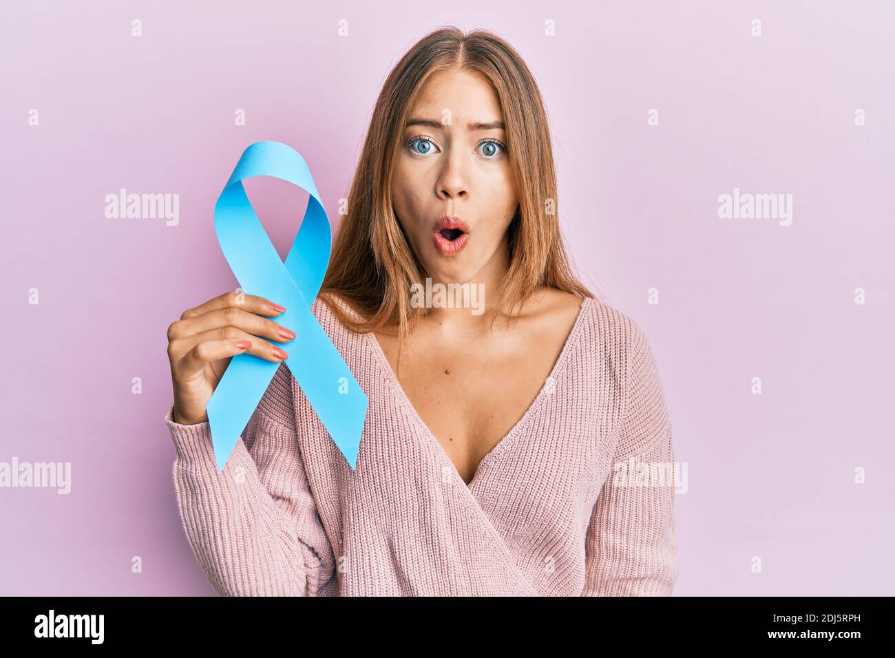 Beautiful young blonde woman holding blue ribbon scared and amazed with ...