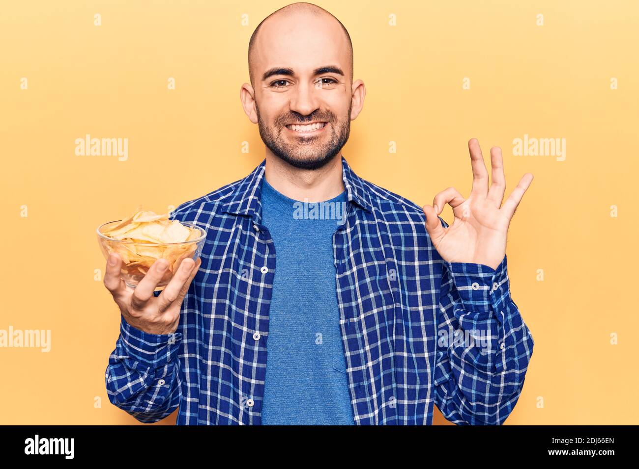 Young handsome bald man holding bowl with potato chips doing ok sign ...