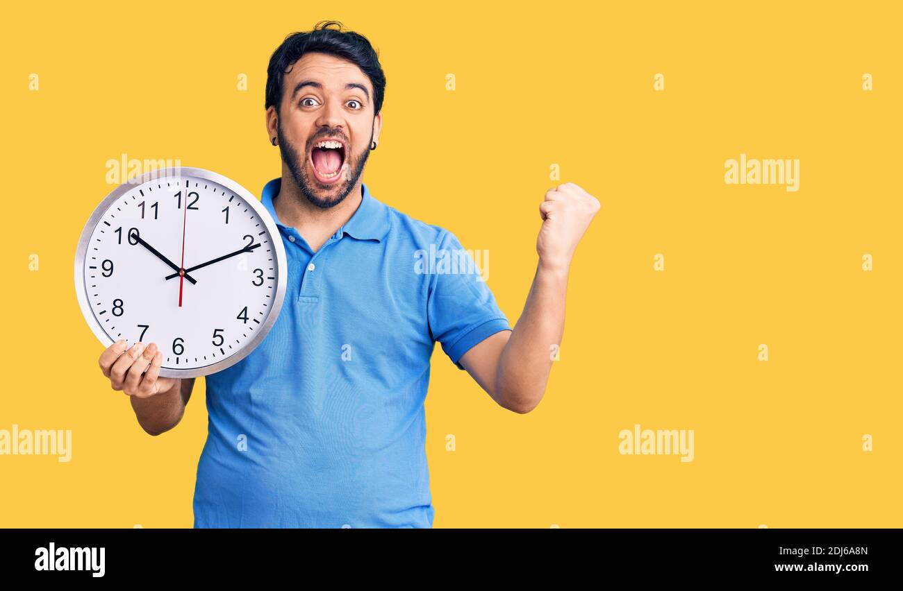 Young hispanic man holding big clock screaming proud, celebrating ...