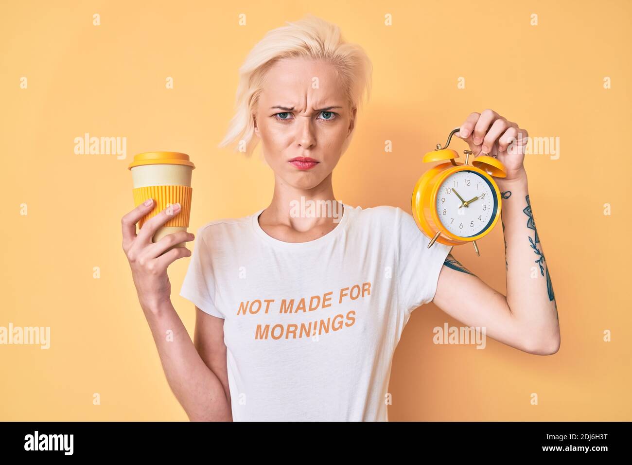 Young blonde woman with tattoo holding alarm clock and take away cup of ...