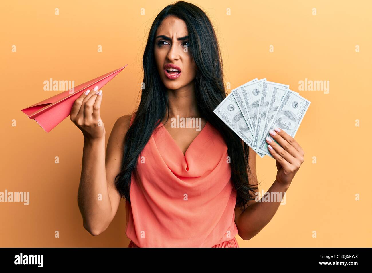 Beautiful hispanic woman holding paper plane and dollars angry and mad ...