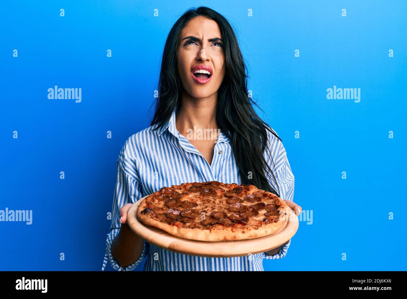 Beautiful hispanic woman holding tasty pepperoni pizza angry and mad ...