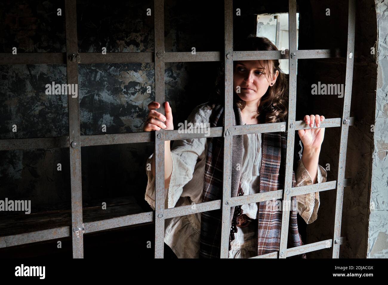 A woman prisoner with a sad smile looks through the bars in a dark ...