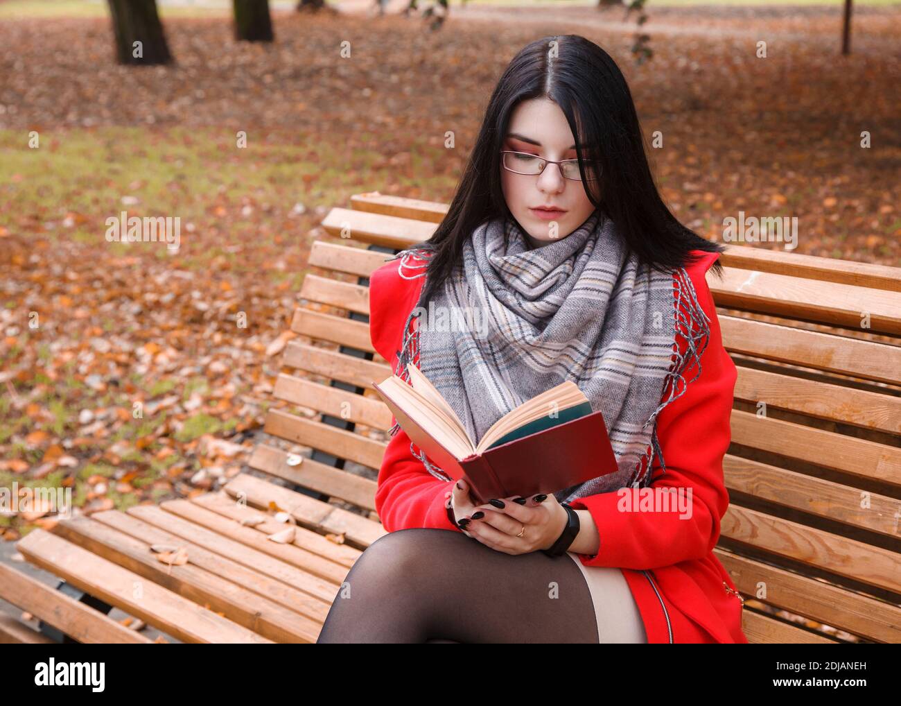 young beautiful girl in red coat reading book while sitting on a park ...