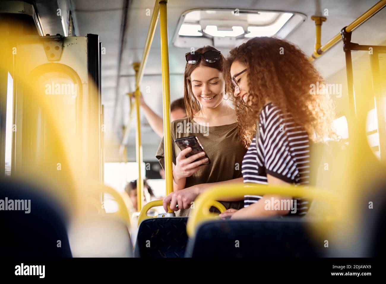 Two cheerful pretty young women are standing in a bus and looking at ...