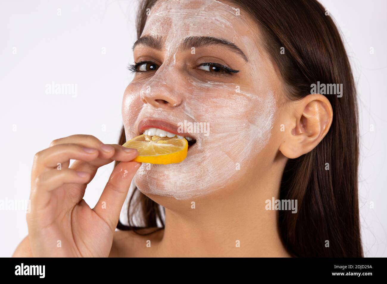 A girl with cream on her face holding slice of lemon in her mouth ...