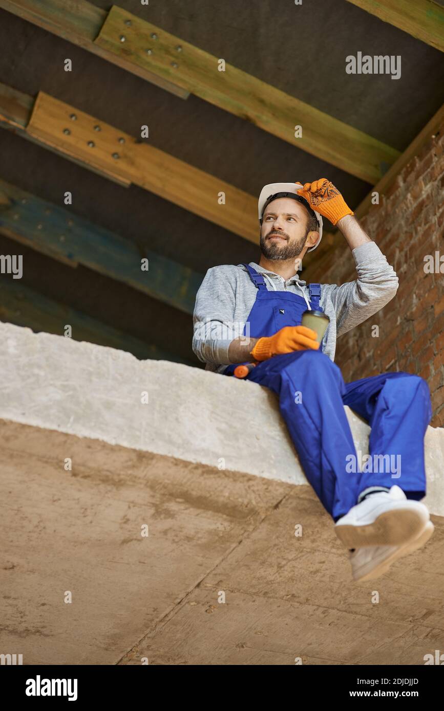 Positive young male builder in blue overalls and hard hat looking ...