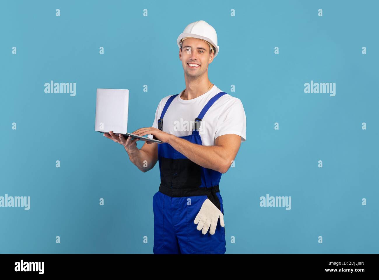 Man in coveralls and hard hat using laptop computer on blue background ...