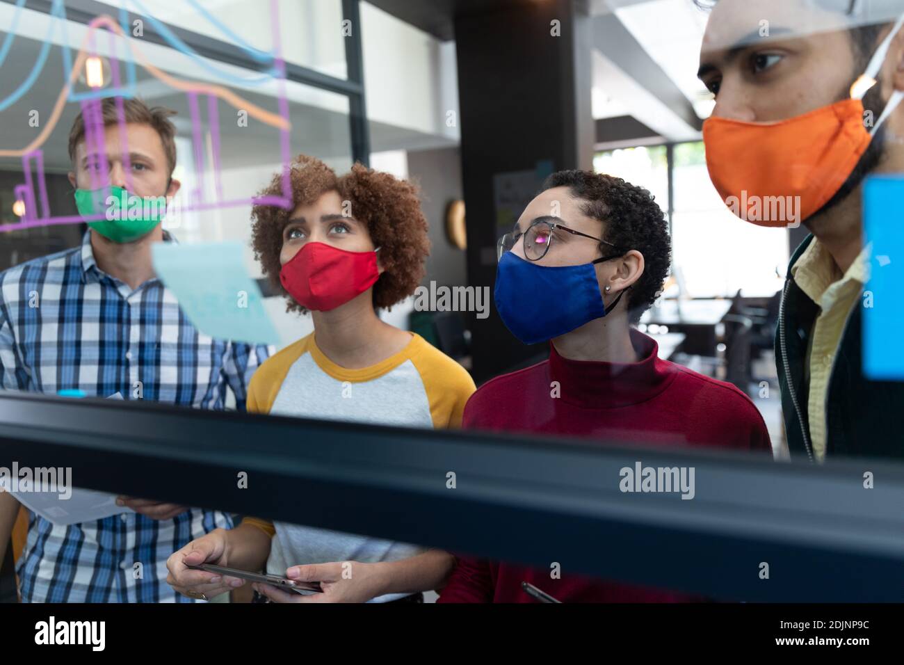Diverse group of colleagues wearing face masks brainstorming at glass ...