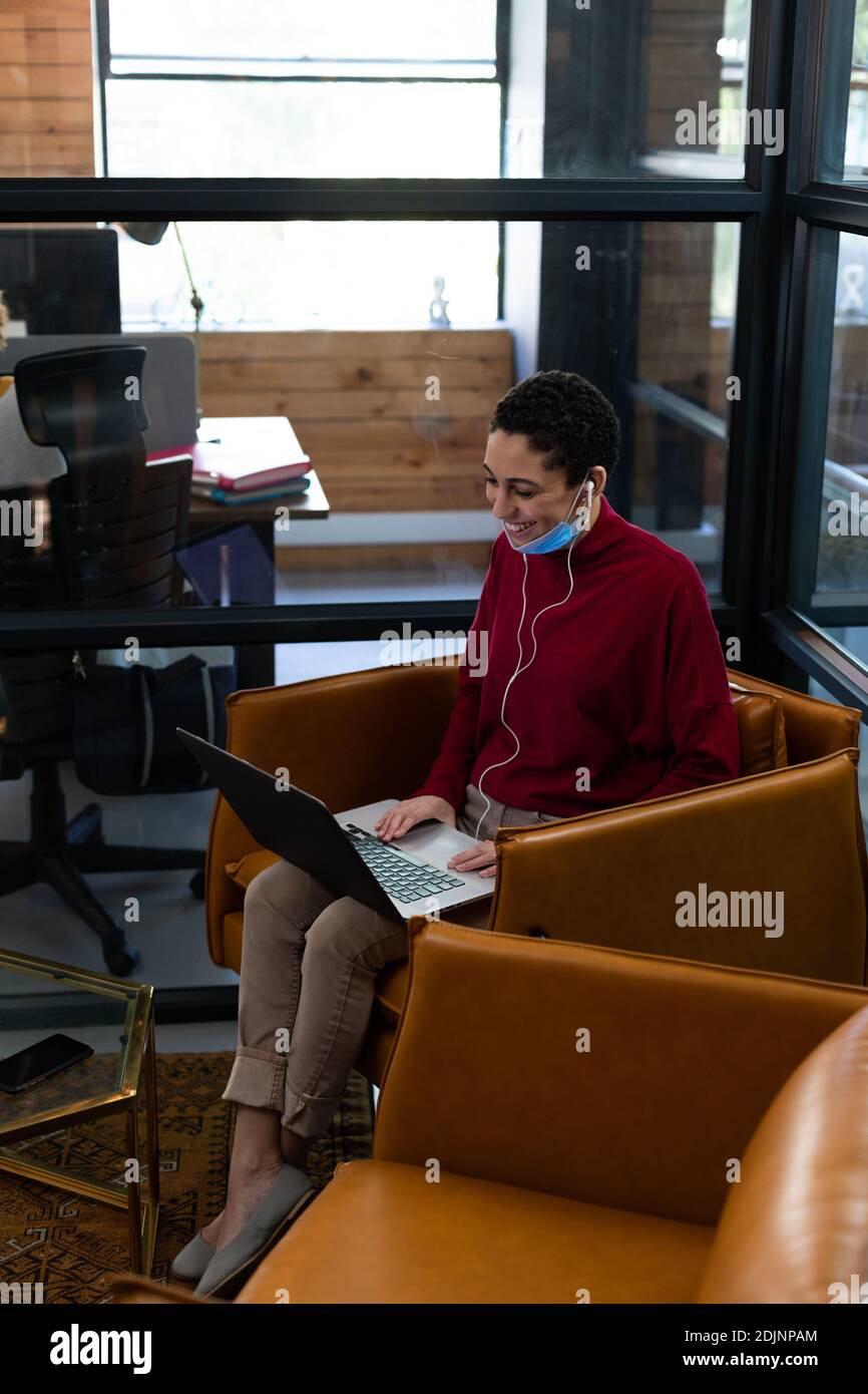 Mixed race woman wearing lowered face mask using laptop and earphones ...