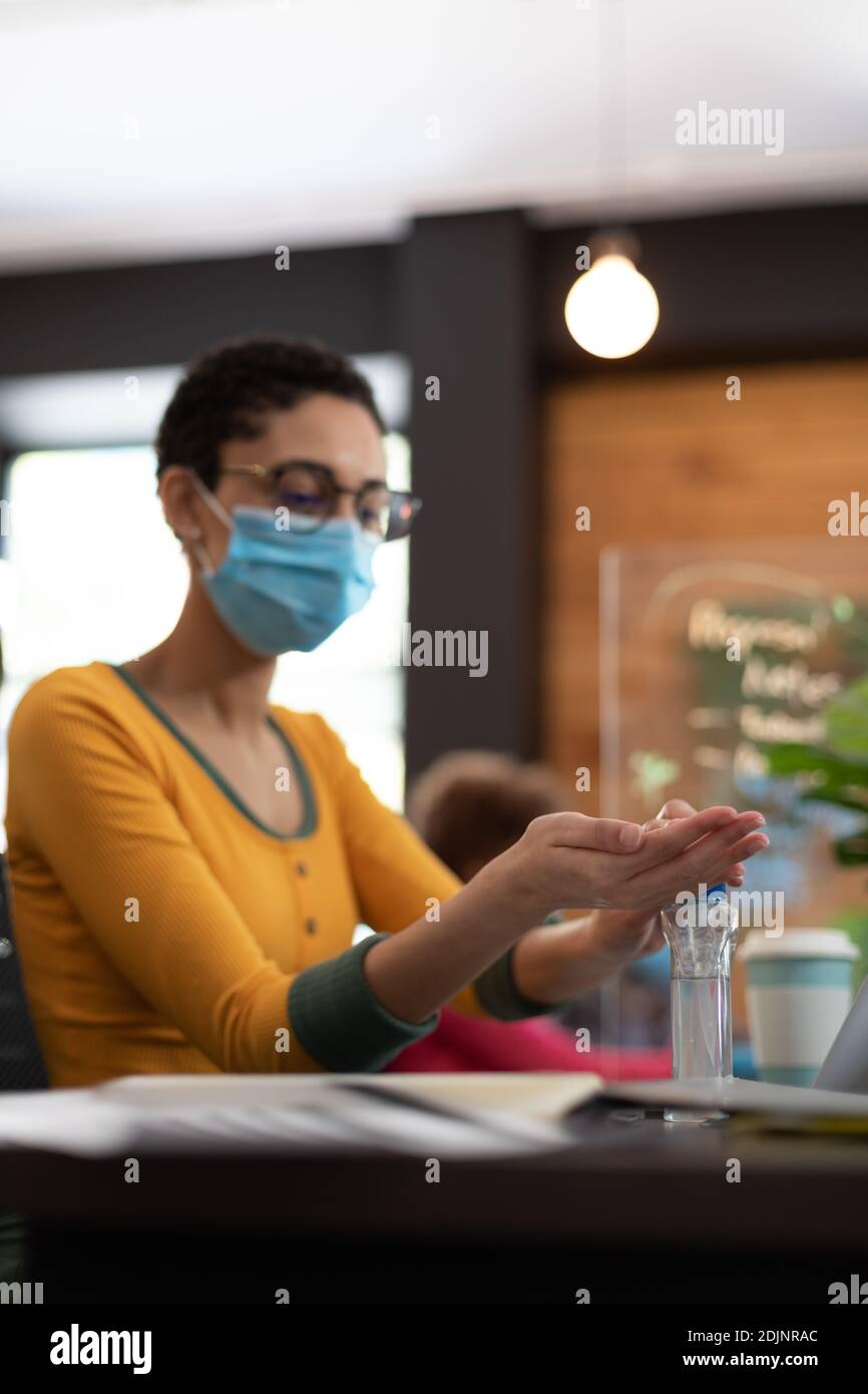 Mixed race woman wearing face mask sanitizing hands at desk in office ...