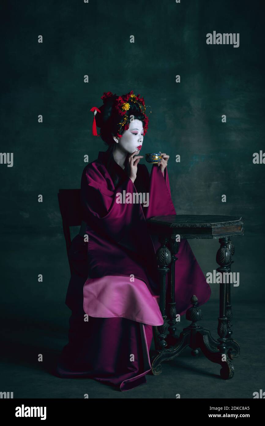 Tea drinking. Young japanese woman as geisha isolated on dark green ...
