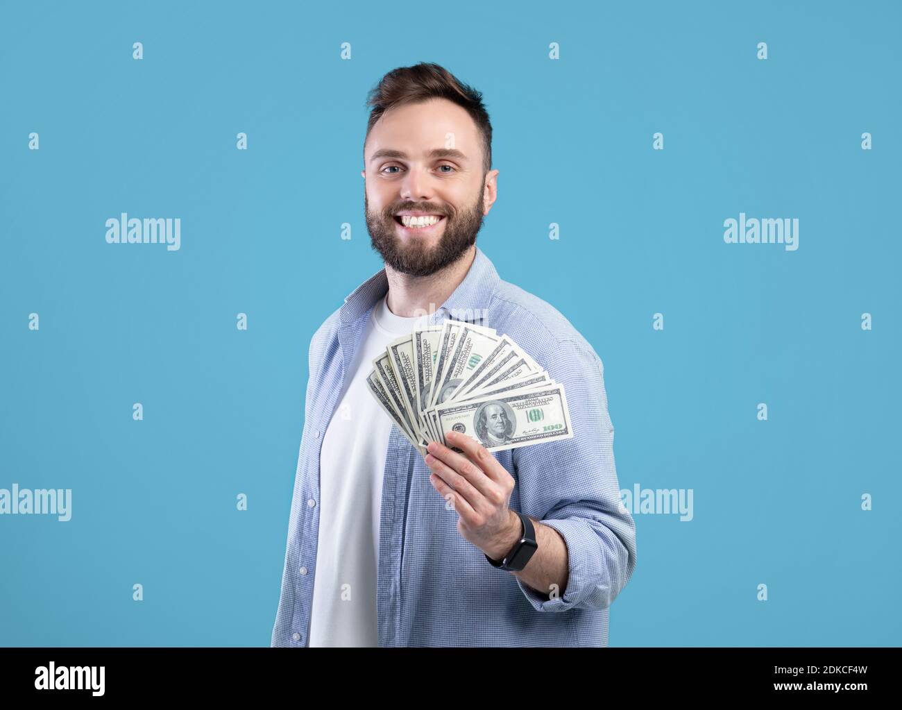 Happy young man posing with lots of money, holding dollar fan over blue ...