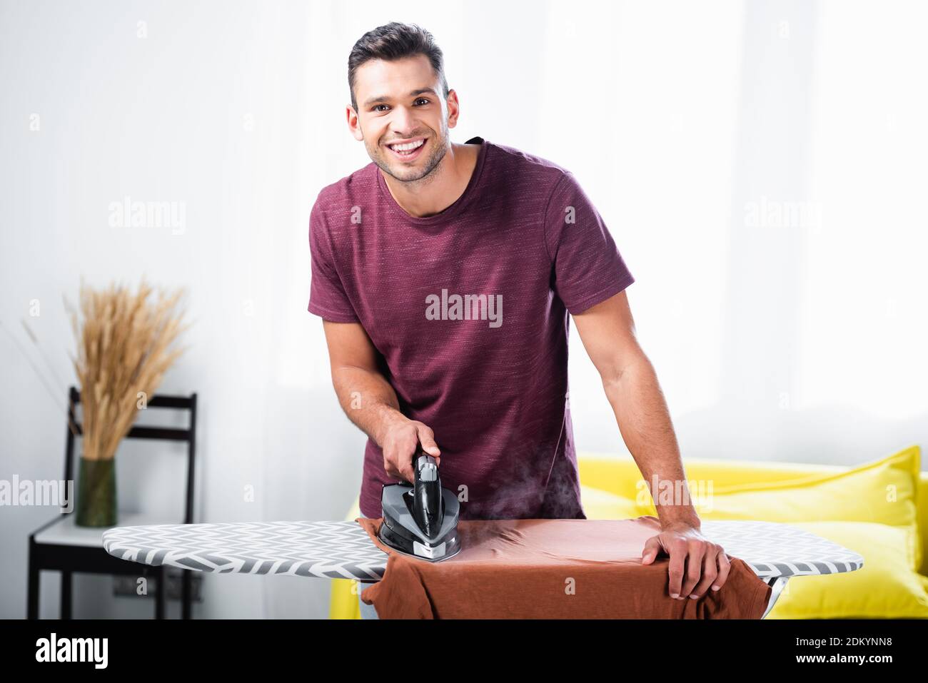 Cheerful man using iron while ironing clothes on board in living room ...