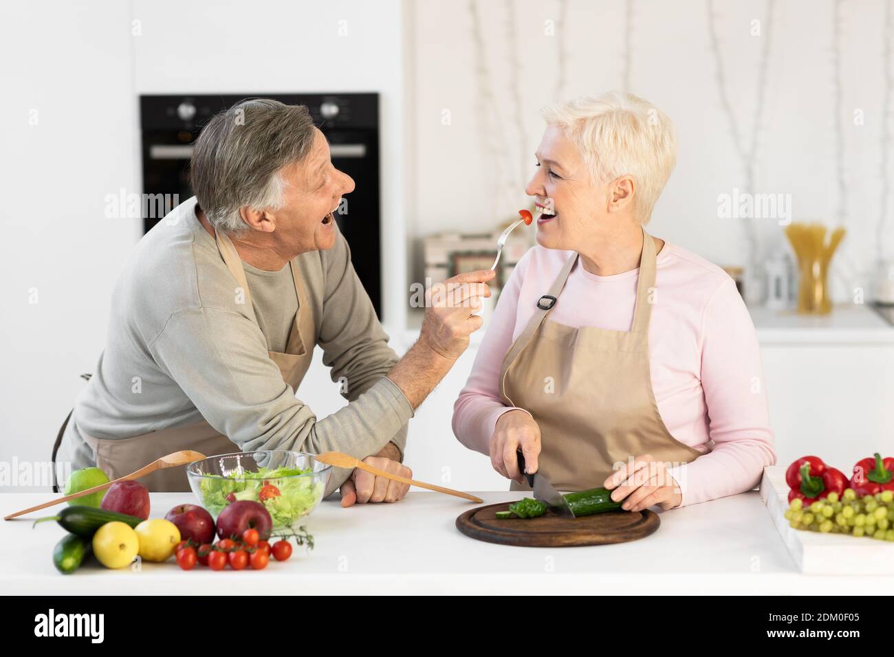 Cheerful Elderly Husband Feeding Wife Tasting Dinner Cooking In Kitchen ...