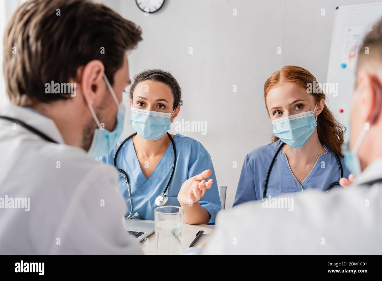 Multicultural nurses in medical masks looking at doctors while sitting ...