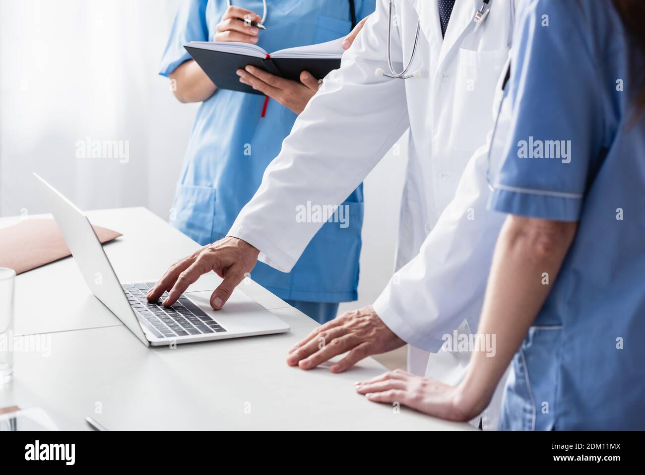 Cropped view of doctor in white coat using laptop near nurses with ...