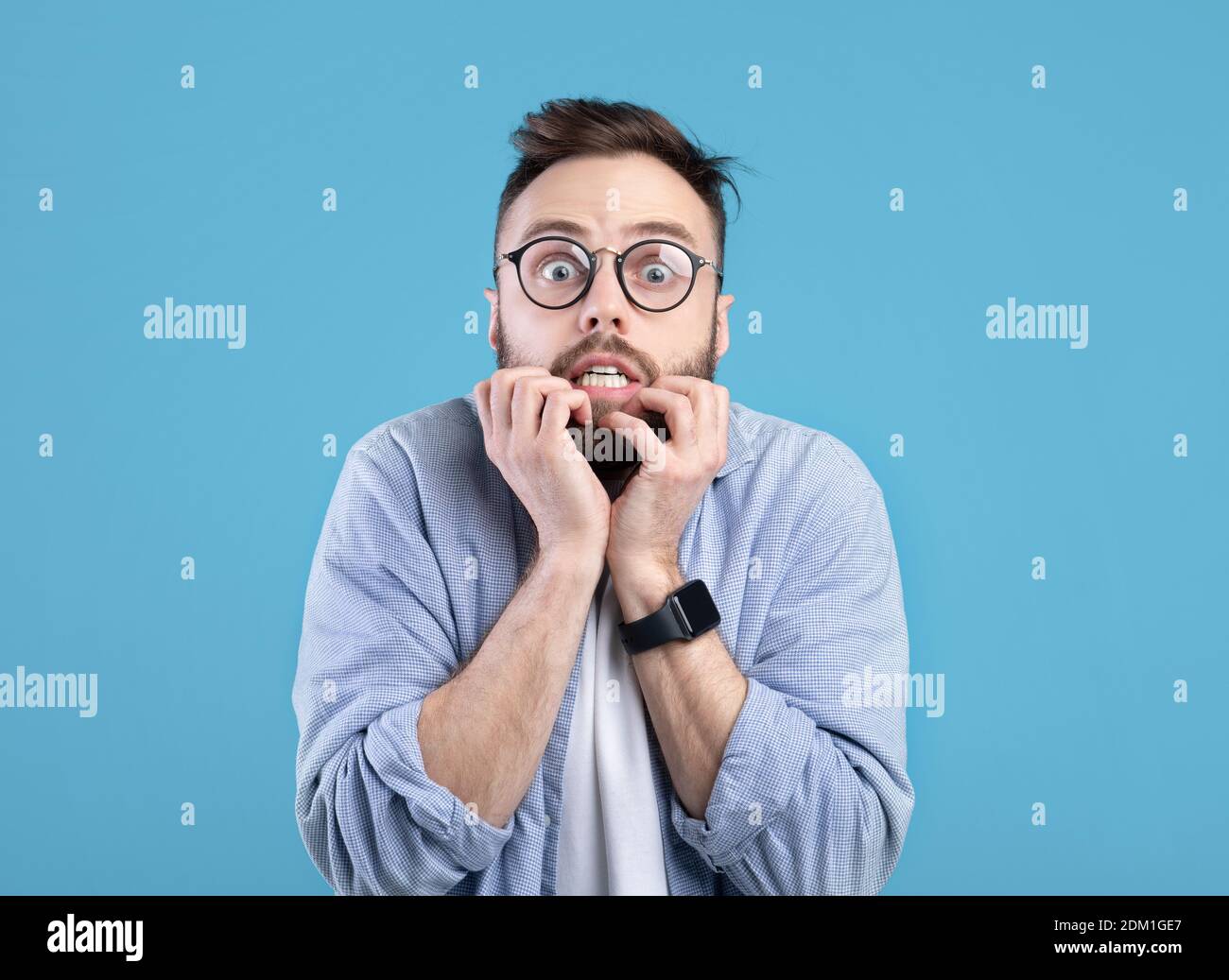 Scared millennial guy biting nails in panic over blue studio background ...