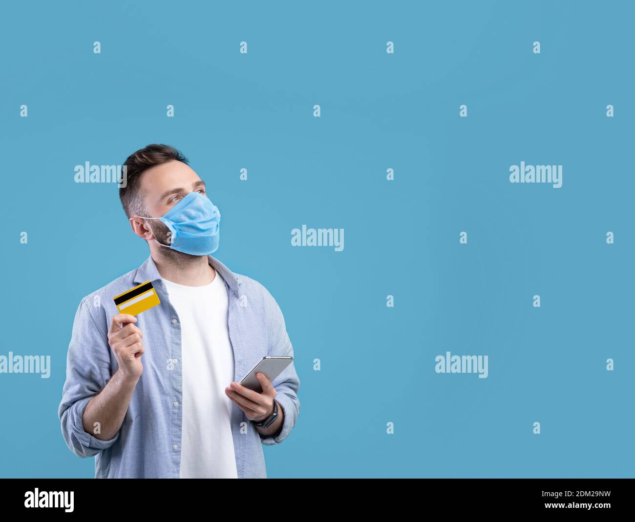 Millennial man in face mask holding smartphone and credit card, looking ...