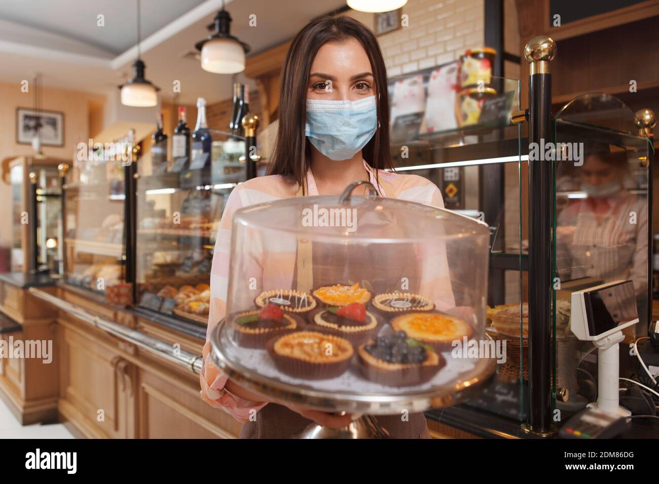 Female baker wearing medical face mask, carrying tray with desserts ...