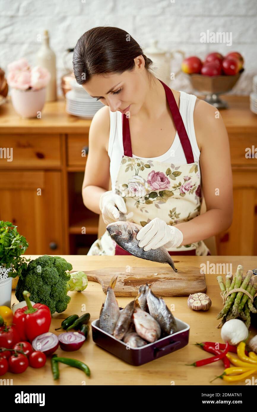 beautiful young woman, brunette prepares fresh fish at a table full of ...