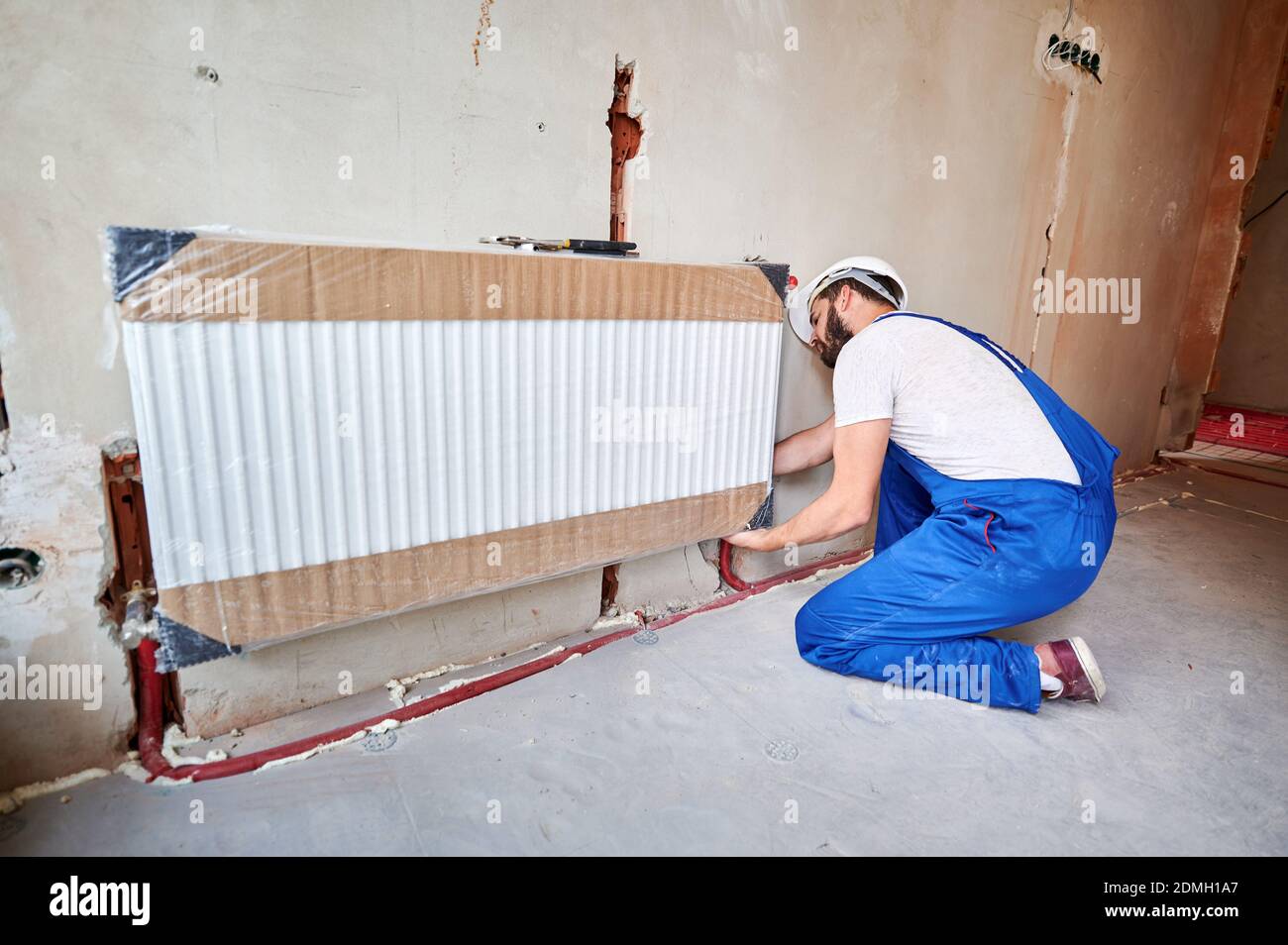 Male plumber in work overalls installing heating radiator in empty room ...