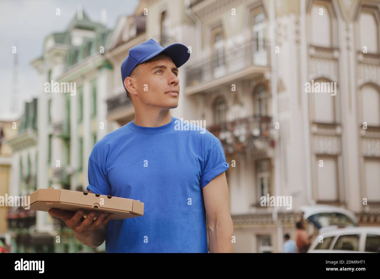 Young pizza deliveryman looking around in the city, carrying pizza box ...