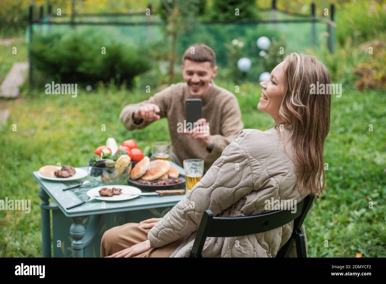 A young man takes photos of his girlfriend on a smartphone during a picnic in nature near their ...