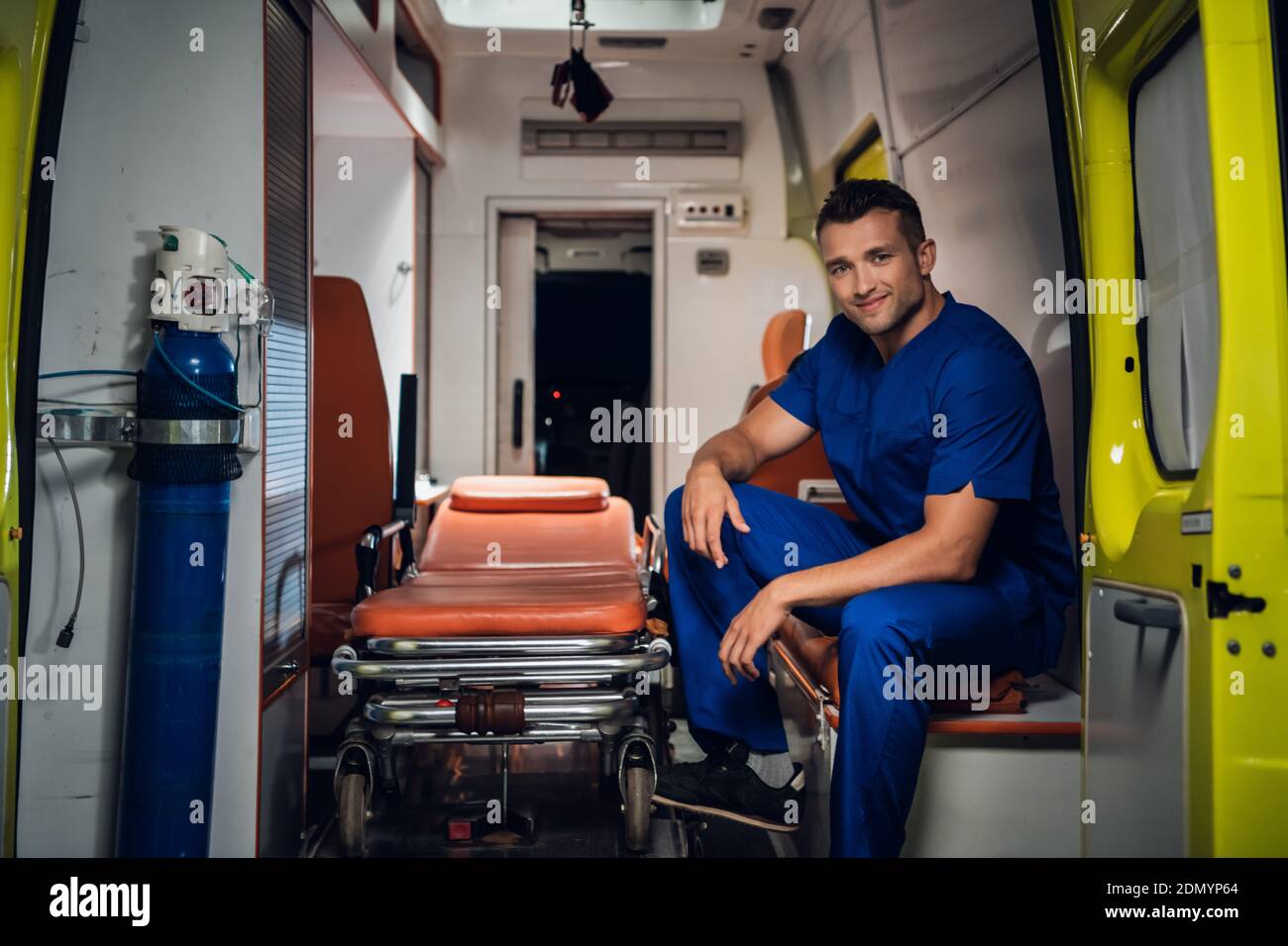 Smiling paramedic in a blue uniform sitting in the back of an ambulance ...