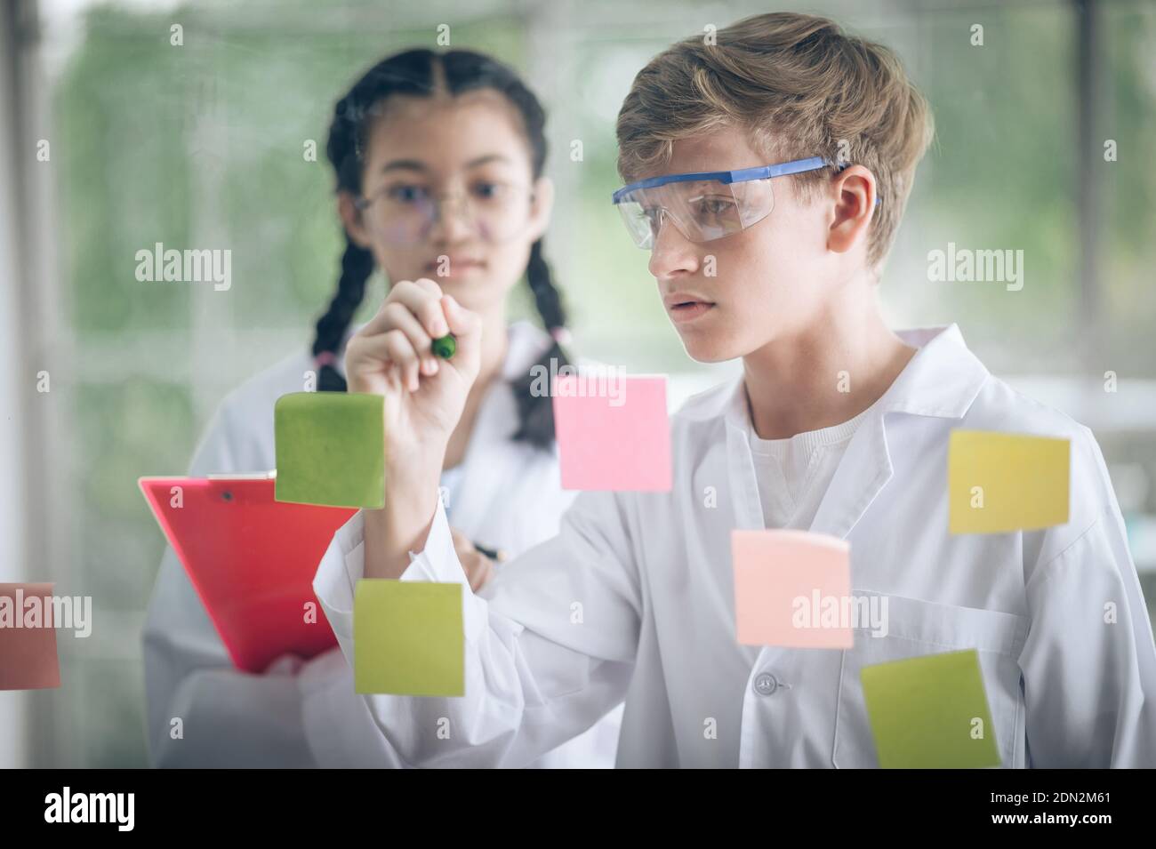 Male Student Writing On Adhesive Notes With Classmate In Background ...