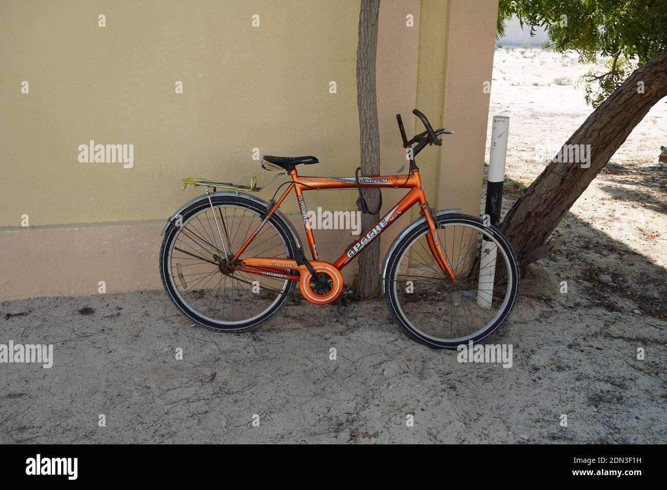 An old orange bicycle is standing on the sand path locked to a tree ...